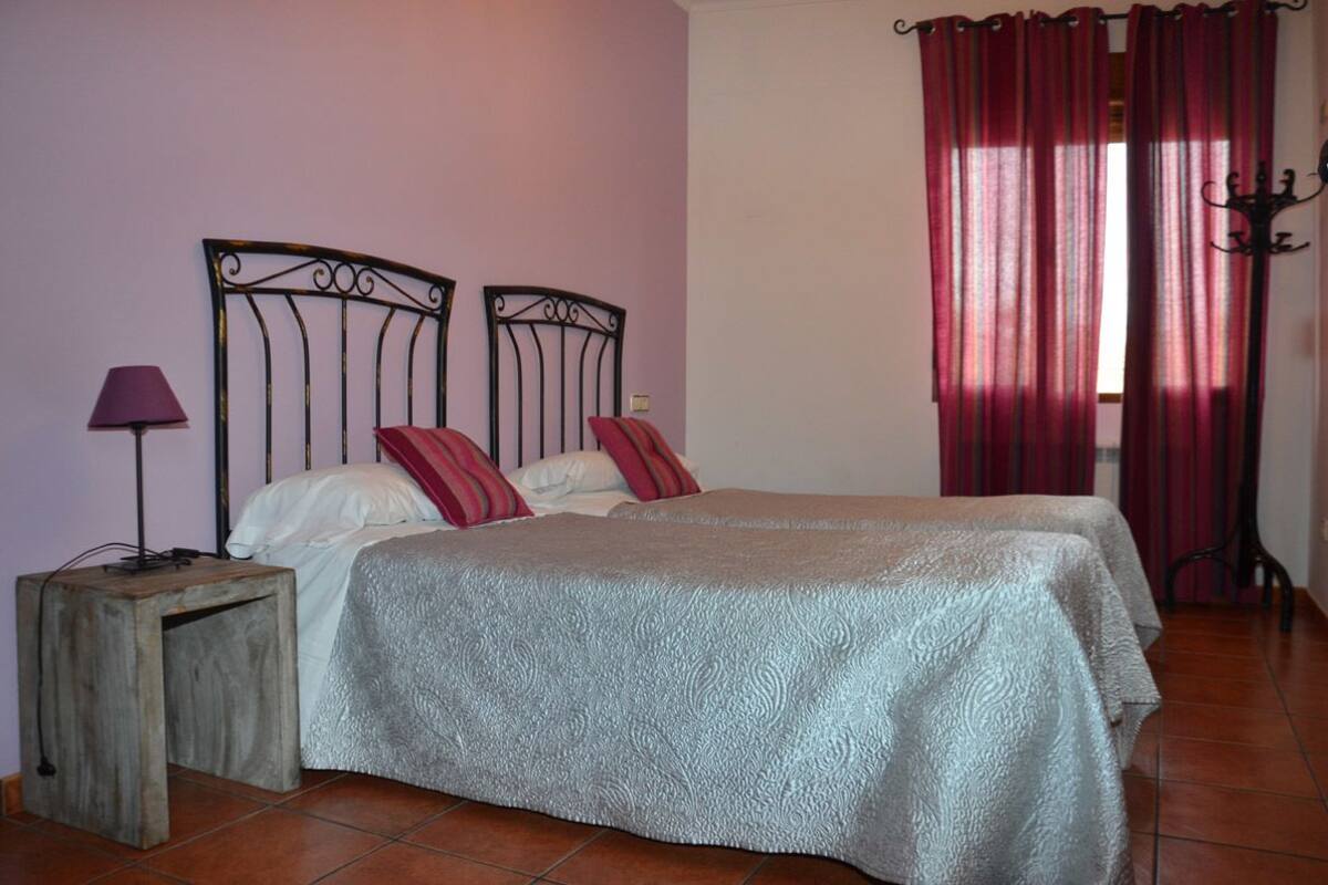 Quarto com duas camas, uma mesa de cabeceira, uma luminária e uma janela com cortinas.