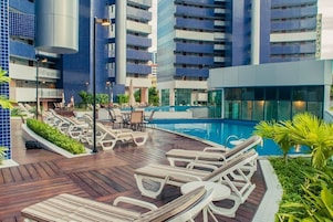 Uma área de piscina com espreguiçadeiras, um deck de madeira e um edifício moderno com varandas.