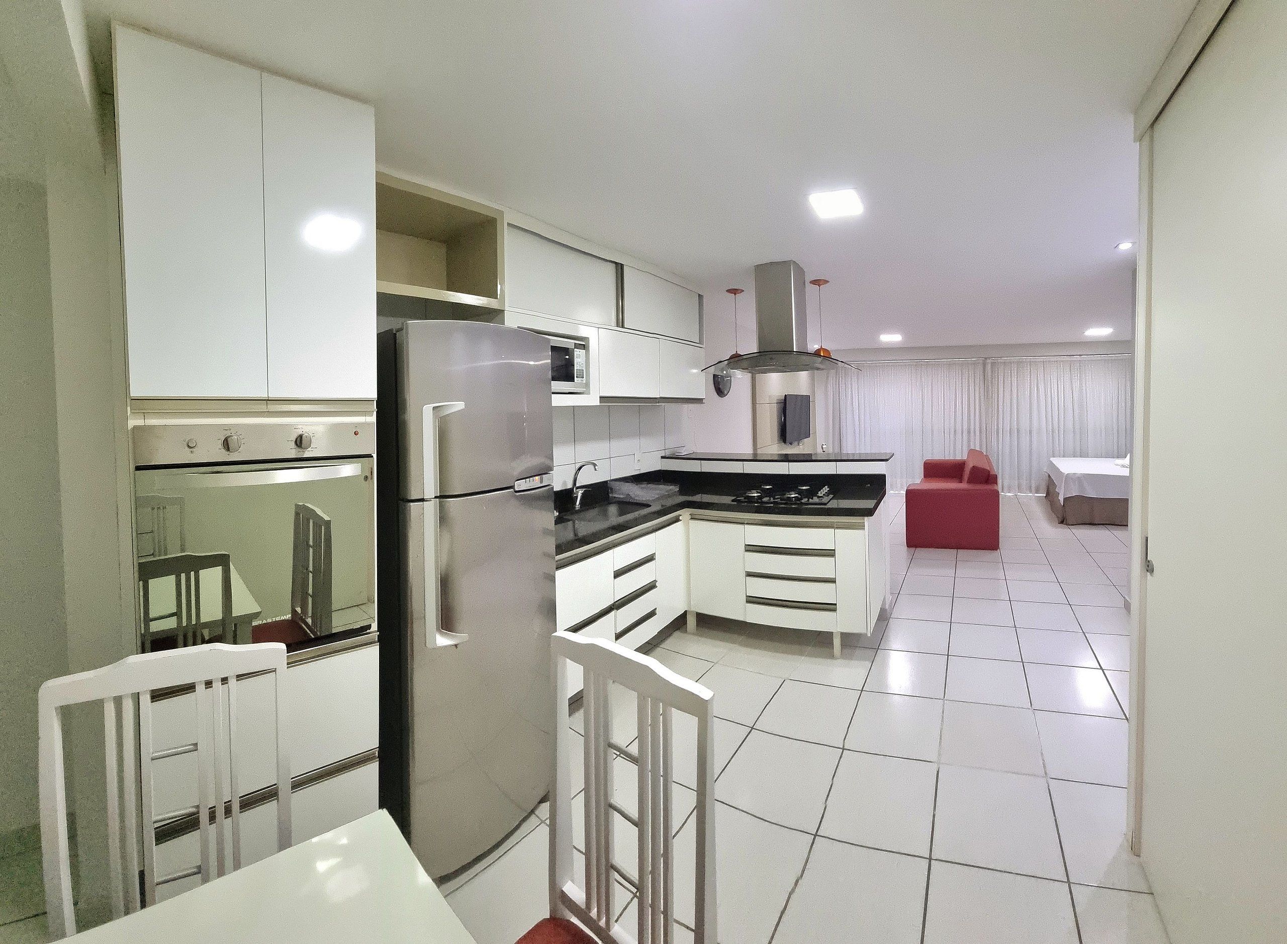 Cozinha moderna com geladeira, fogão e micro-ondas. Há uma mesa de jantar com cadeiras e um sofá vermelho na sala de estar.