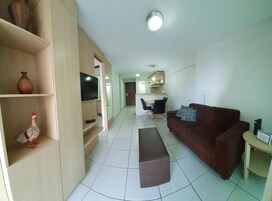 Uma sala de estar com um sofá marrom, uma televisão em uma estante, uma área de jantar com mesa e cadeiras, e uma cozinha ao fundo.