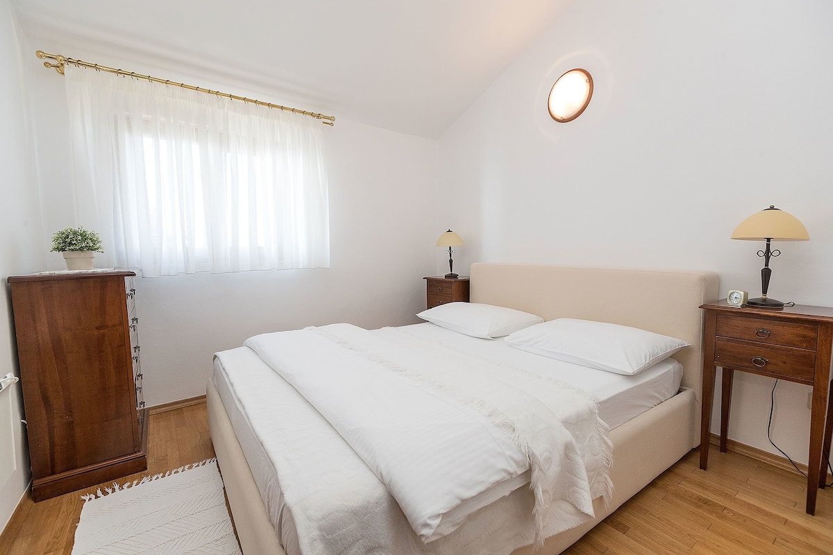 Quarto com cama, mesa de cabeceira, cômoda, abajur e janela com cortinas.