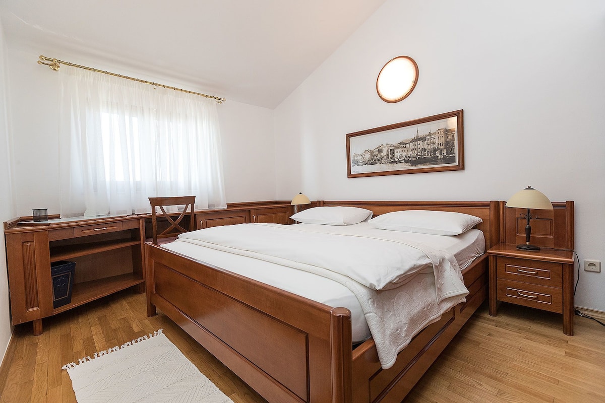 Quarto com cama de madeira, mesas de cabeceira, escrivaninha e janela com cortinas leves.
