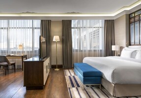 Quarto de hotel com uma cama grande, uma escrivaninha, uma cadeira, uma TV e vista da cidade através de amplas janelas.