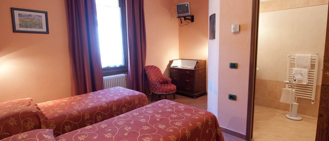 Quarto de hotel com duas camas, uma escrivaninha, uma cadeira, um radiador, uma televisão e um porta-toalhas.