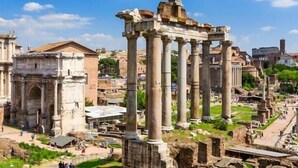 Ruínas antigas com colunas e um grande arco, rodeadas por vegetação e outras estruturas históricas.