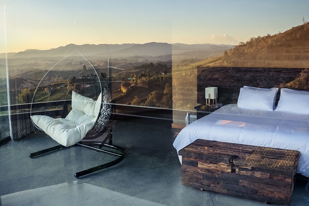 Quarto com cama, um baú de madeira e uma cadeira suspensa com vista para as montanhas.