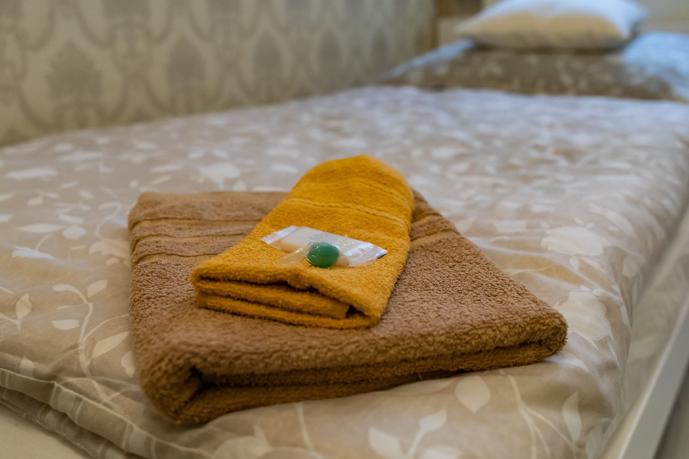 Toalha marrom dobrada com um objeto verde sobre a cama, com roupa de cama estampada.