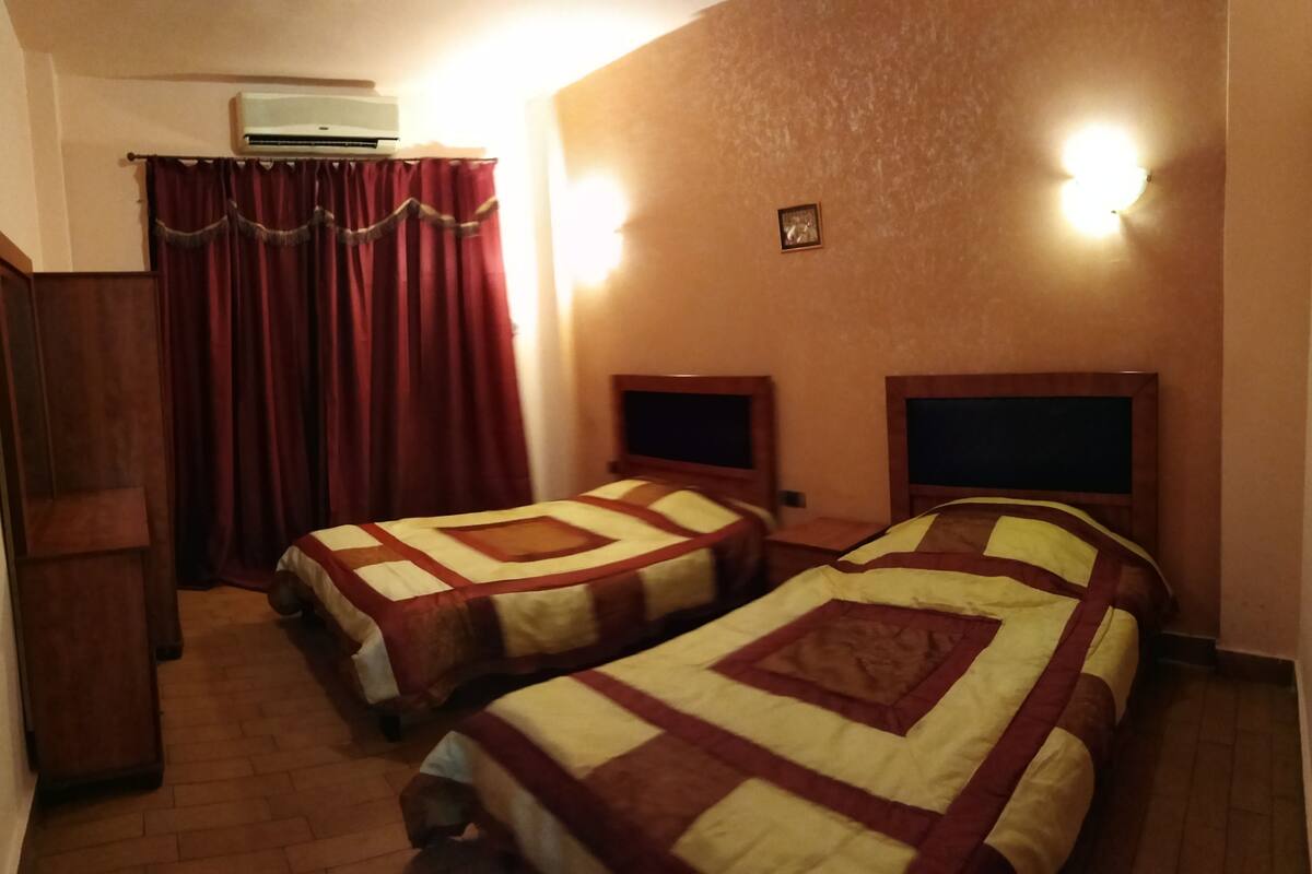 Quarto com duas camas, um guarda-roupa de madeira, cortina vermelha, aparelho de ar condicionado e luminária na parede.