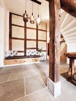 Interior rústico com vigas de madeira aparentes, piso de ladrilho de pedra, um banco de madeira com almofadas e iluminação pendente.