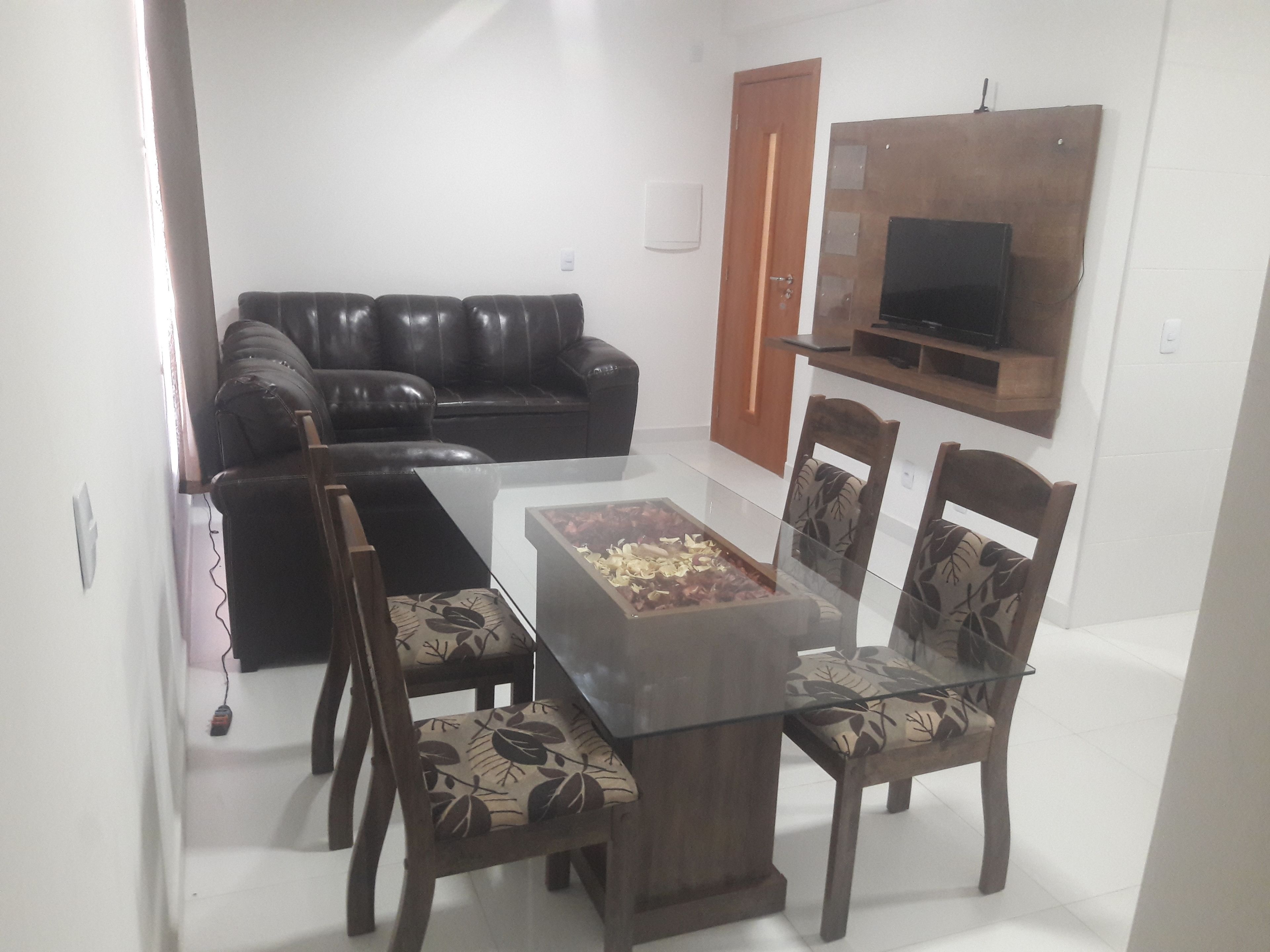 Uma sala de estar com uma mesa de jantar de vidro, quatro cadeiras, um sofá de couro preto, uma TV montada na parede e um armário de madeira abaixo da TV.