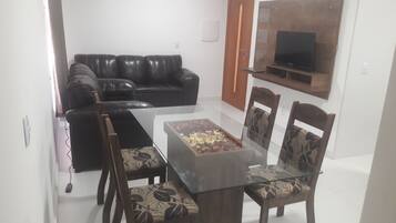 Uma sala de estar com uma mesa de jantar de vidro, quatro cadeiras, um sofá de couro preto, uma TV montada na parede e um armário de madeira abaixo da TV.