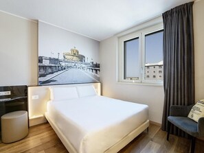 Quarto de hotel com cama, escrivaninha, cadeira, janela com cortinas e uma obra de arte pendurada na parede.
