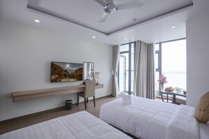 Quarto de hotel com duas camas, uma escrivaninha, uma cadeira, um quadro e vista para o mar.