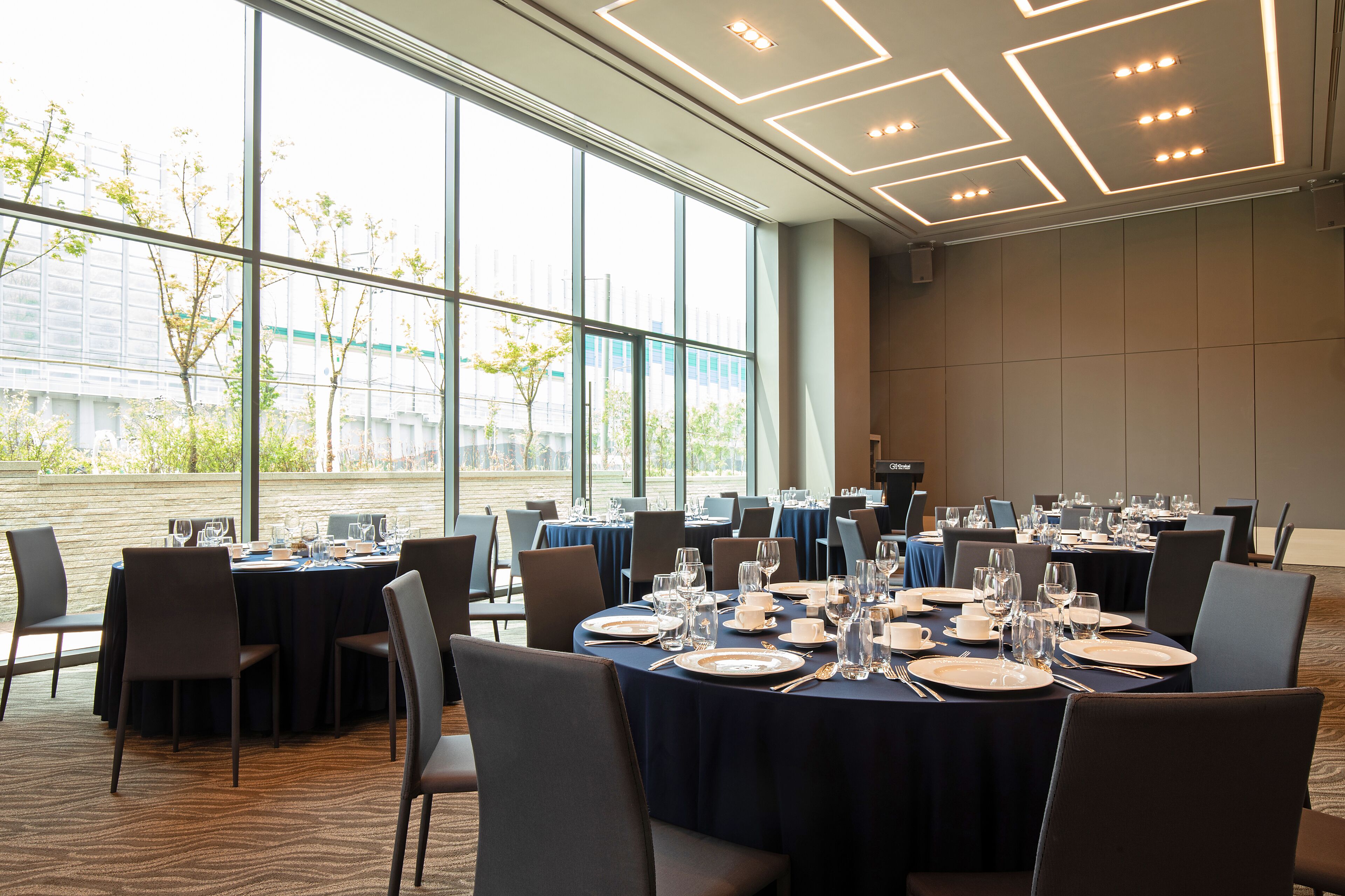 Uma sala de conferências com mesas redondas preparadas para uma reunião, grandes janelas com vista para árvores e edifícios, e um design de teto moderno.