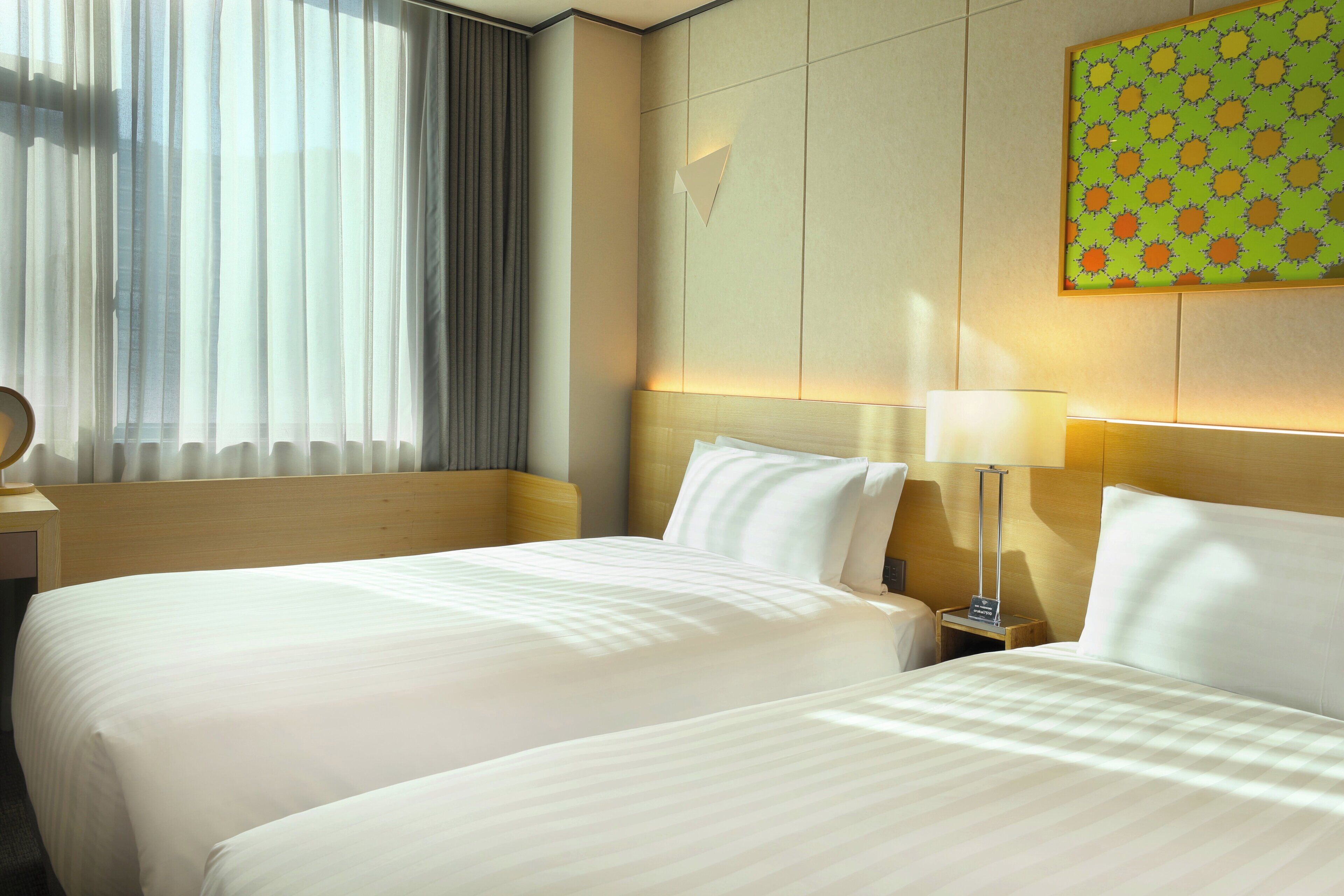 Quarto de hotel com duas camas, uma mesa de cabeceira com abajur, uma janela com cortinas e uma obra de arte na parede.