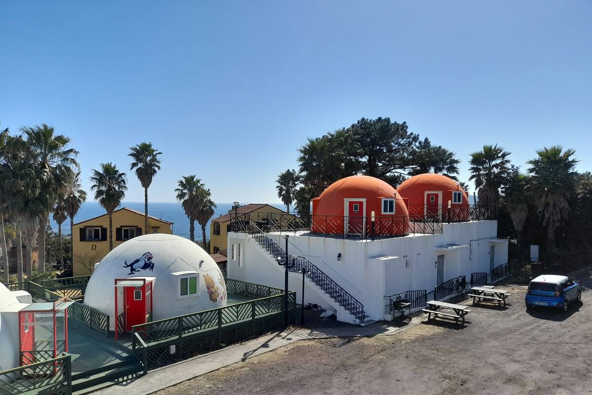 Um edifício com uma estrutura em forma de cúpula e uma porta vermelha, ao lado de uma escadaria que leva a um edifício com telhado plano e duas cúpulas vermelhas.