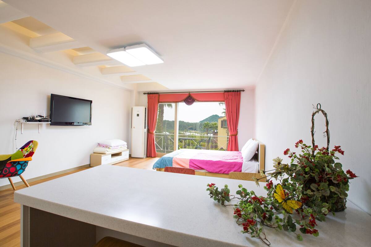 Quarto de hotel com uma cama, uma televisão, uma varanda com cortinas vermelhas e uma cadeira colorida.