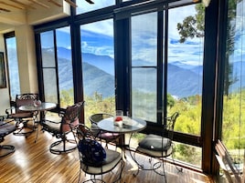 Quarto com amplas janelas oferecendo vista para as montanhas, piso de madeira, cadeiras e mesas com estrutura de metal.