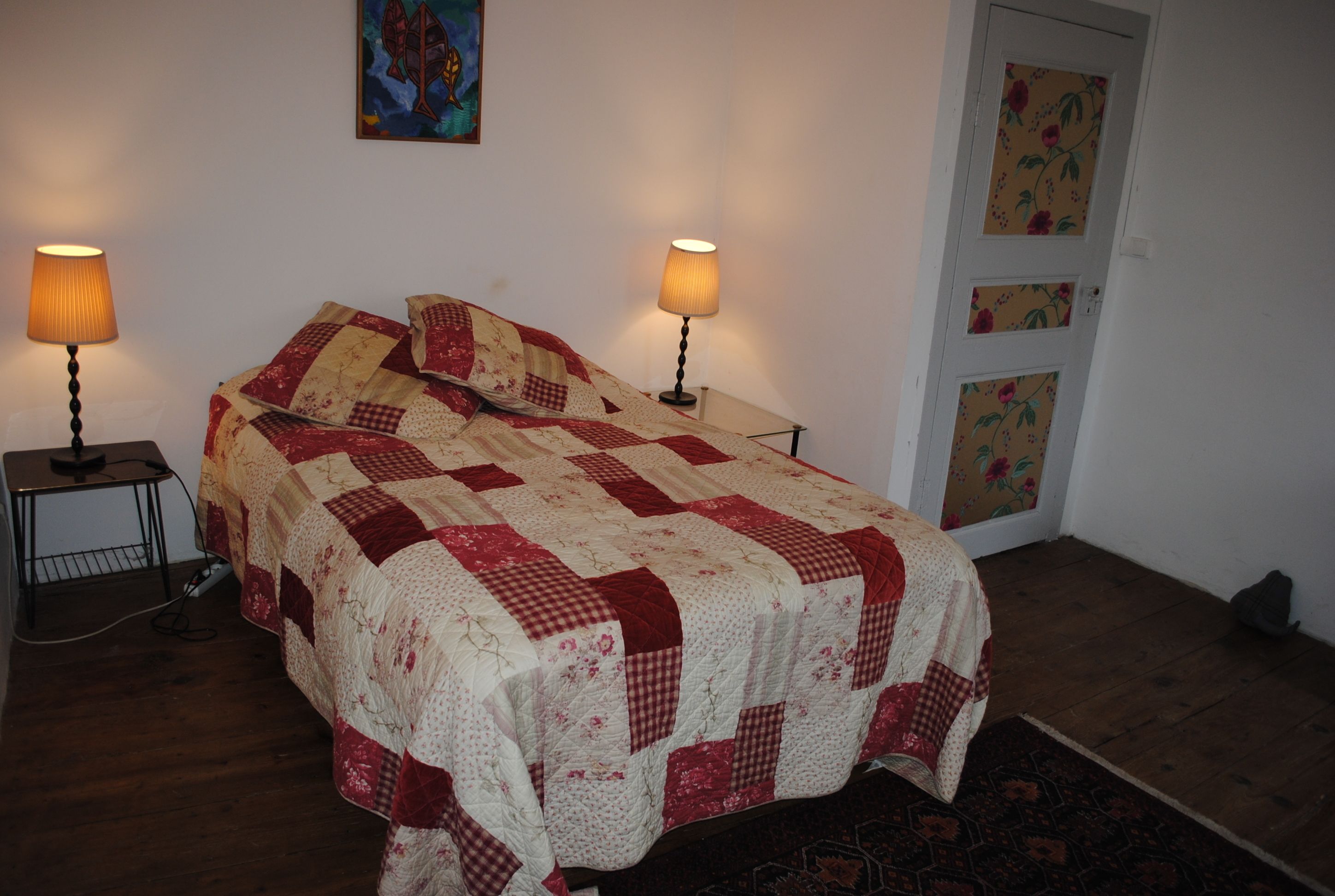 Quarto com cama coberta por um colcha de patchwork, duas luminárias de cabeceira, um quadro na parede e uma porta com papel de parede floral.
