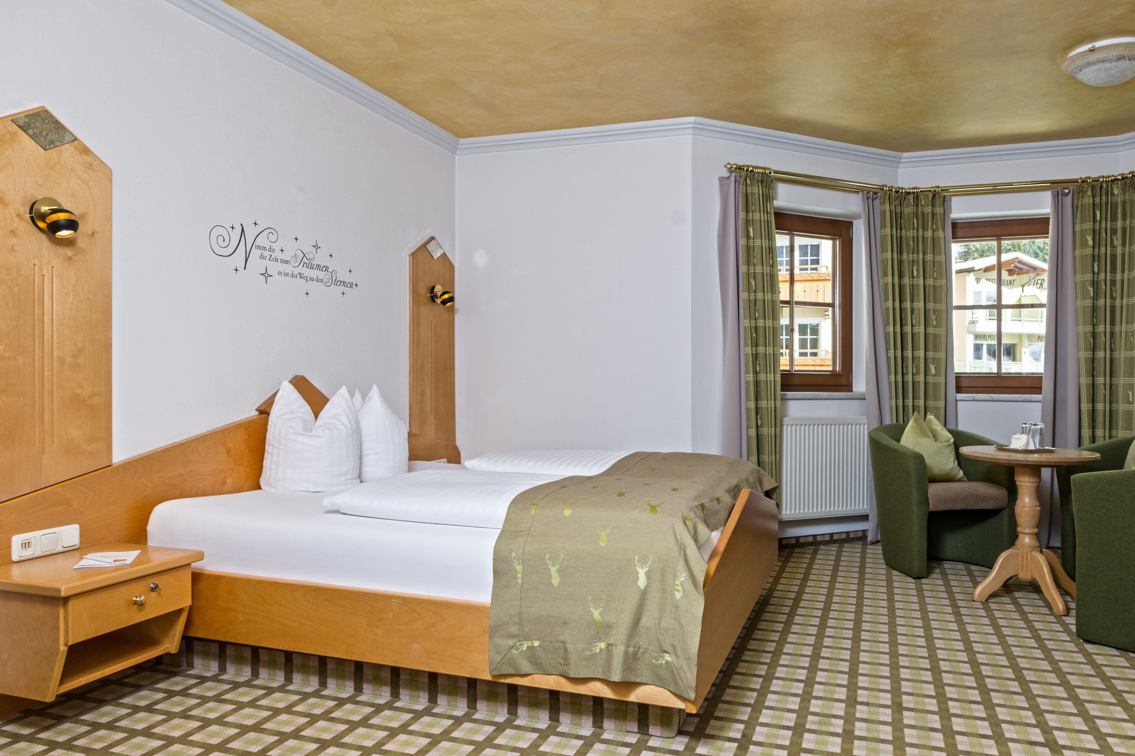 Quarto de hotel com cama de madeira, uma pequena mesa de cabeceira, uma poltrona verde, uma mesa redonda de madeira e uma janela com cortinas.