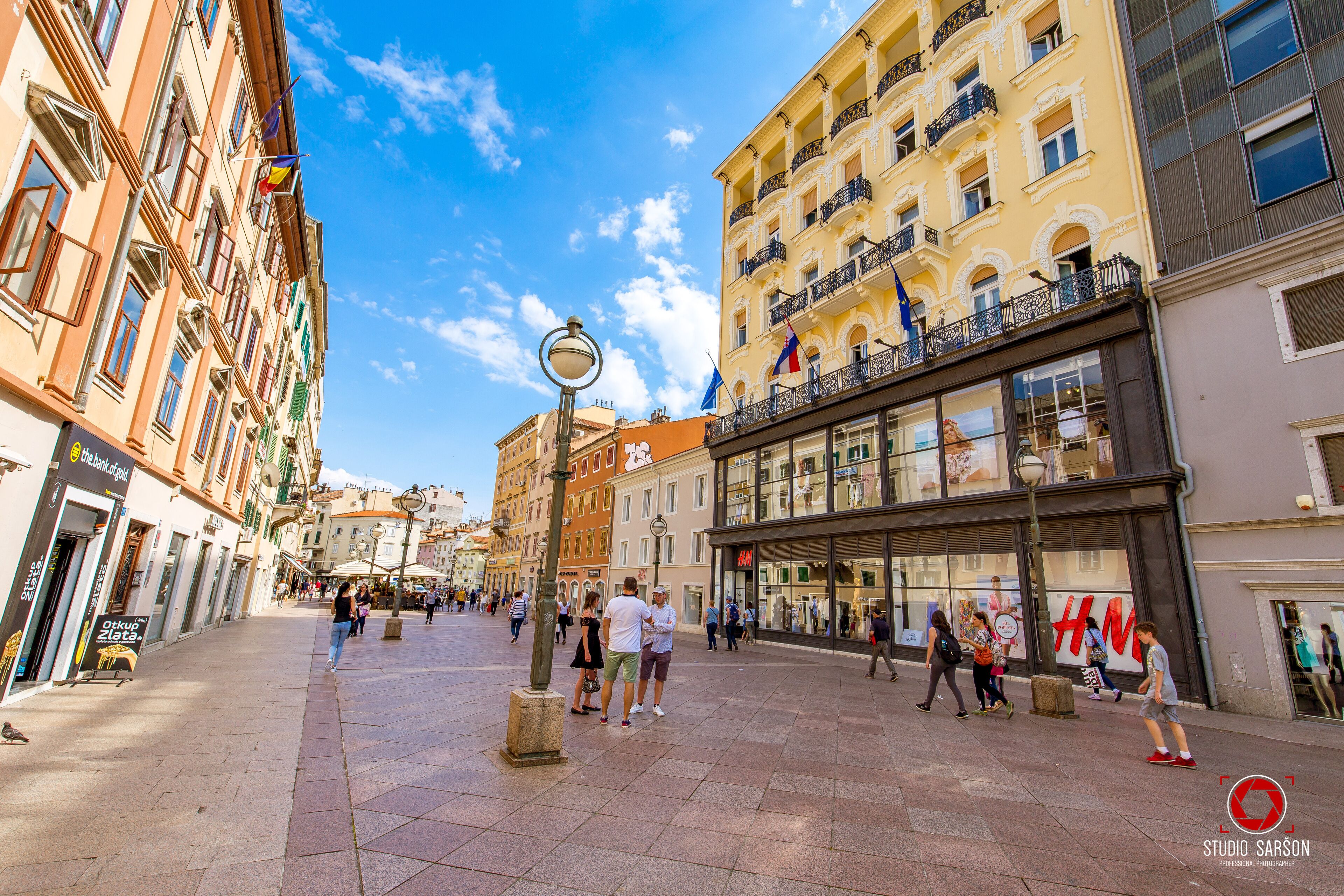 Uma rua destinada a pedestres, ladeada por edifícios históricos, alguns com sacadas e janelas amplas, sob um céu limpo.