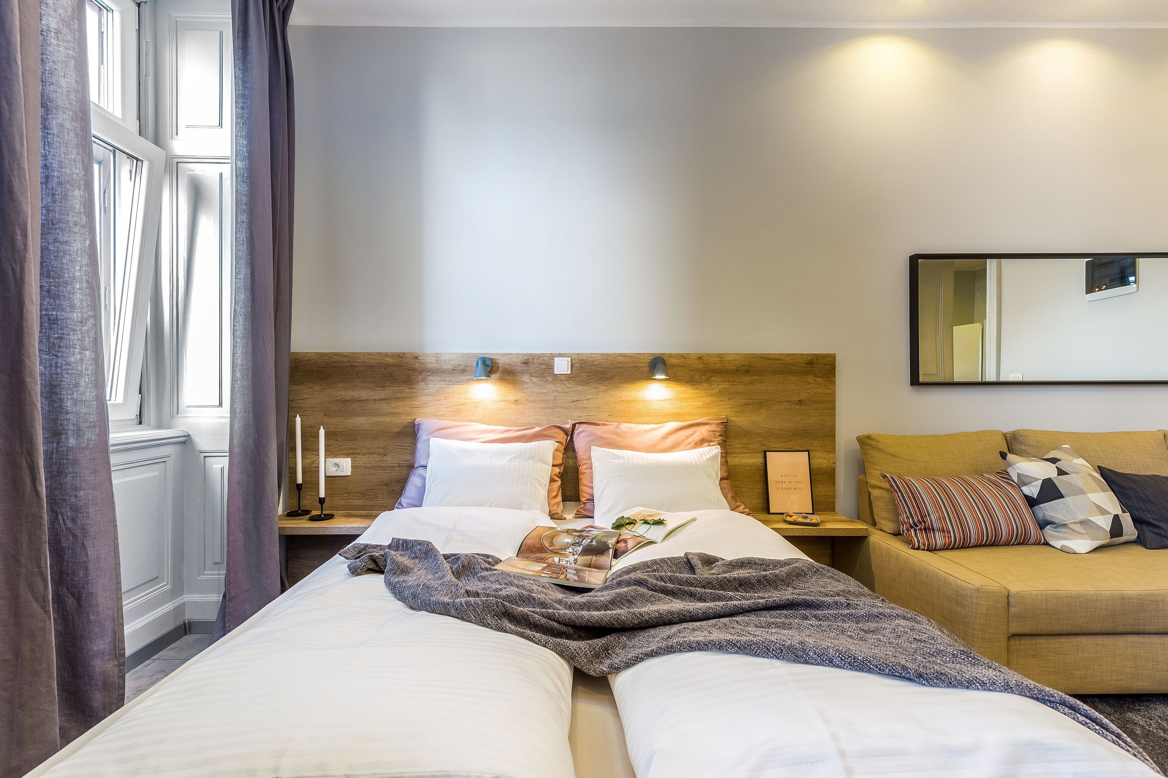 Quarto de hotel com duas camas, cabeceira de madeira, luminárias laterais, mesa de cabeceira, sofá e espelho.
