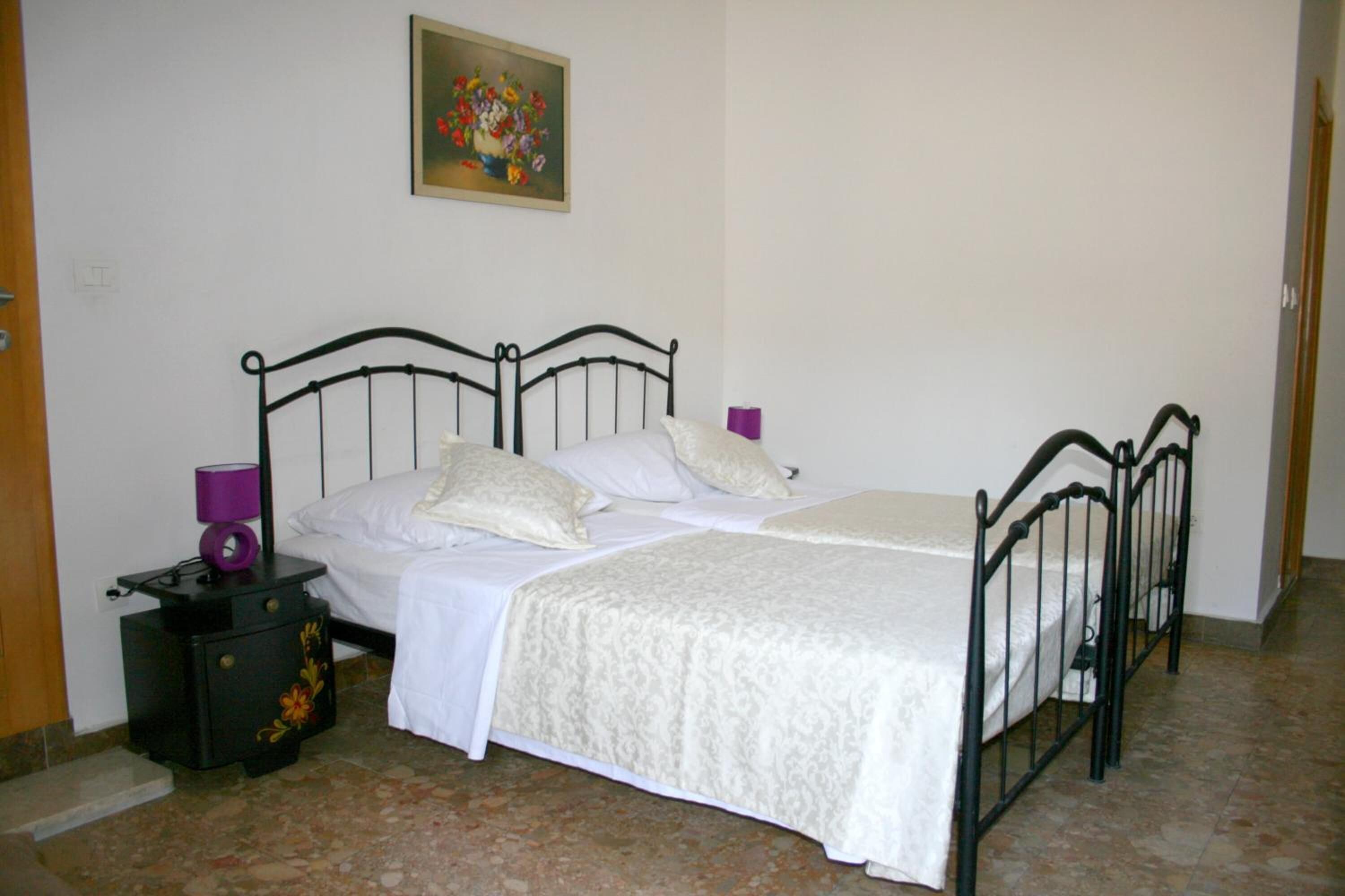 Uma cama bem arrumada com colcha e almofadas brancas, uma cabeceira de metal preto, uma pequena mesa de cabeceira com um abajur roxo e um quadro com flores na parede.