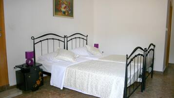 Uma cama bem arrumada com colcha e almofadas brancas, uma cabeceira de metal preto, uma pequena mesa de cabeceira com um abajur roxo e um quadro com flores na parede.