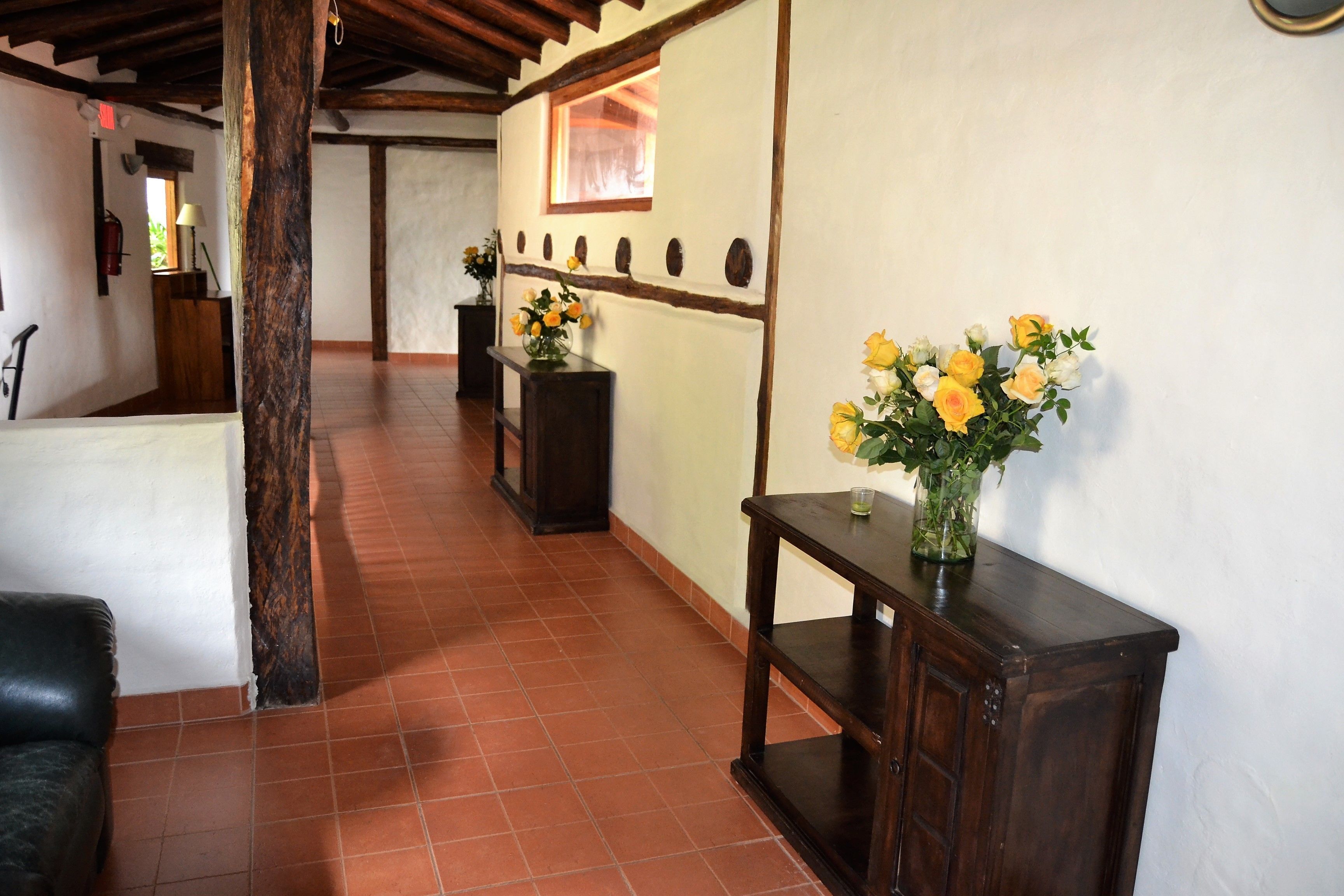 Um corredor com piso de madeira, uma mesa escura de madeira com um vaso de flores e um extintor de incêndio na parede.