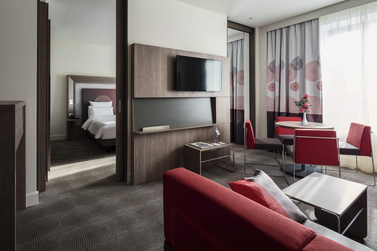 Quarto de hotel com cama, televisão fixada na parede, escrivaninha com cadeira, sofá vermelho e área de refeições com mesa e cadeiras.