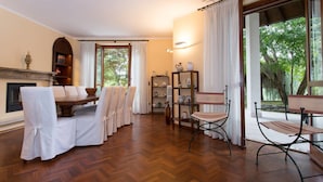 Um quarto amplo com piso de madeira em padrão herringbone, uma mesa de jantar com cadeiras, uma lareira, uma estante de livros e grandes janelas com cortinas.