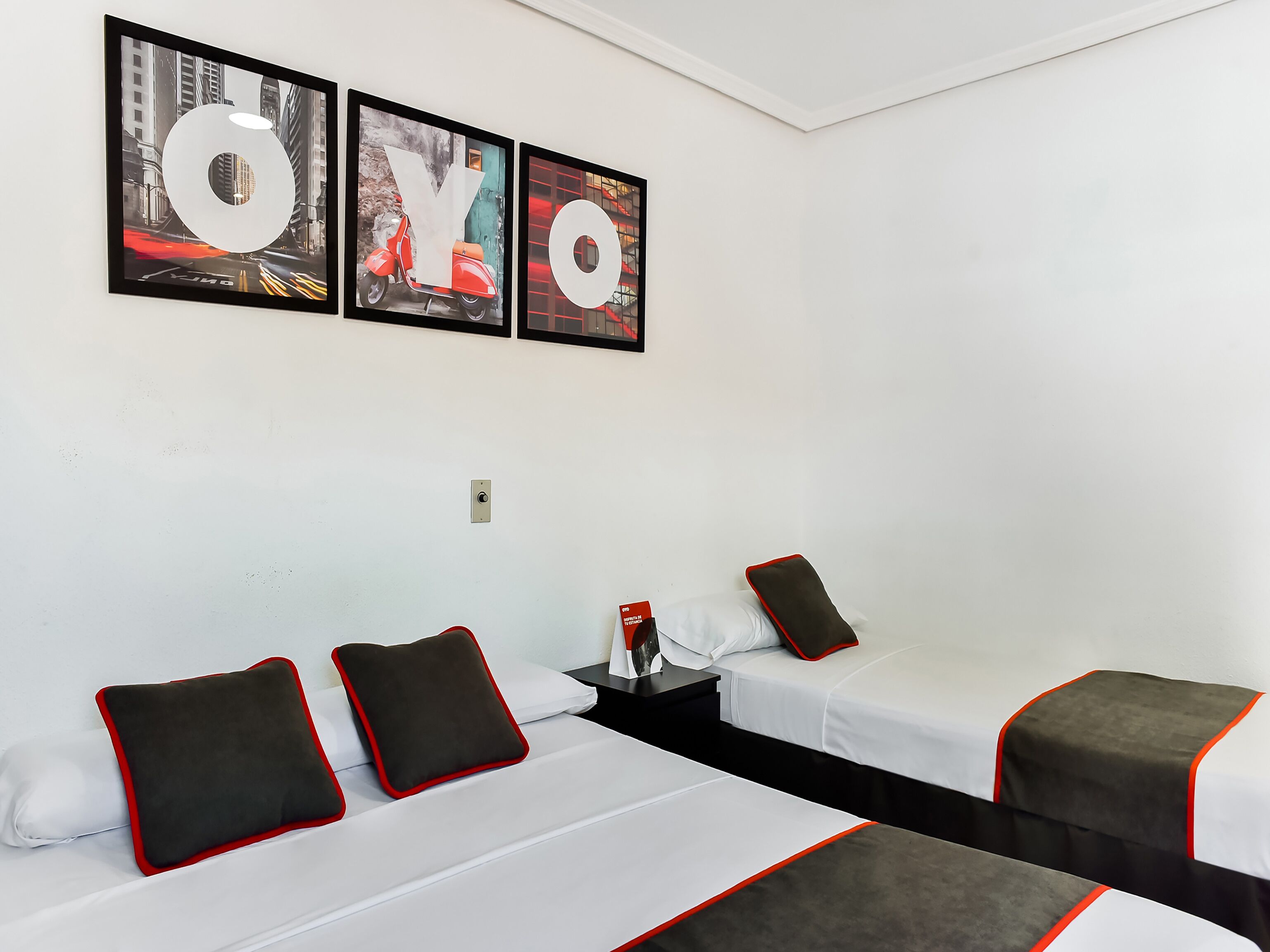 Quarto de hotel com duas camas, cada uma com roupa de cama branca e almofadas com detalhes vermelhos, e uma mesa de cabeceira com abajur vermelho e branco.