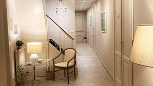 Um corredor com piso em padrão herringbone, uma cadeira, uma mesa com abajur e uma escada.