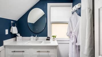 Casa de banho moderna com uma parede de destaque azul escuro, um lavatório branco com um espelho grande, uma janela com persianas e um roupão branco pendurado numa barra de toalhas.