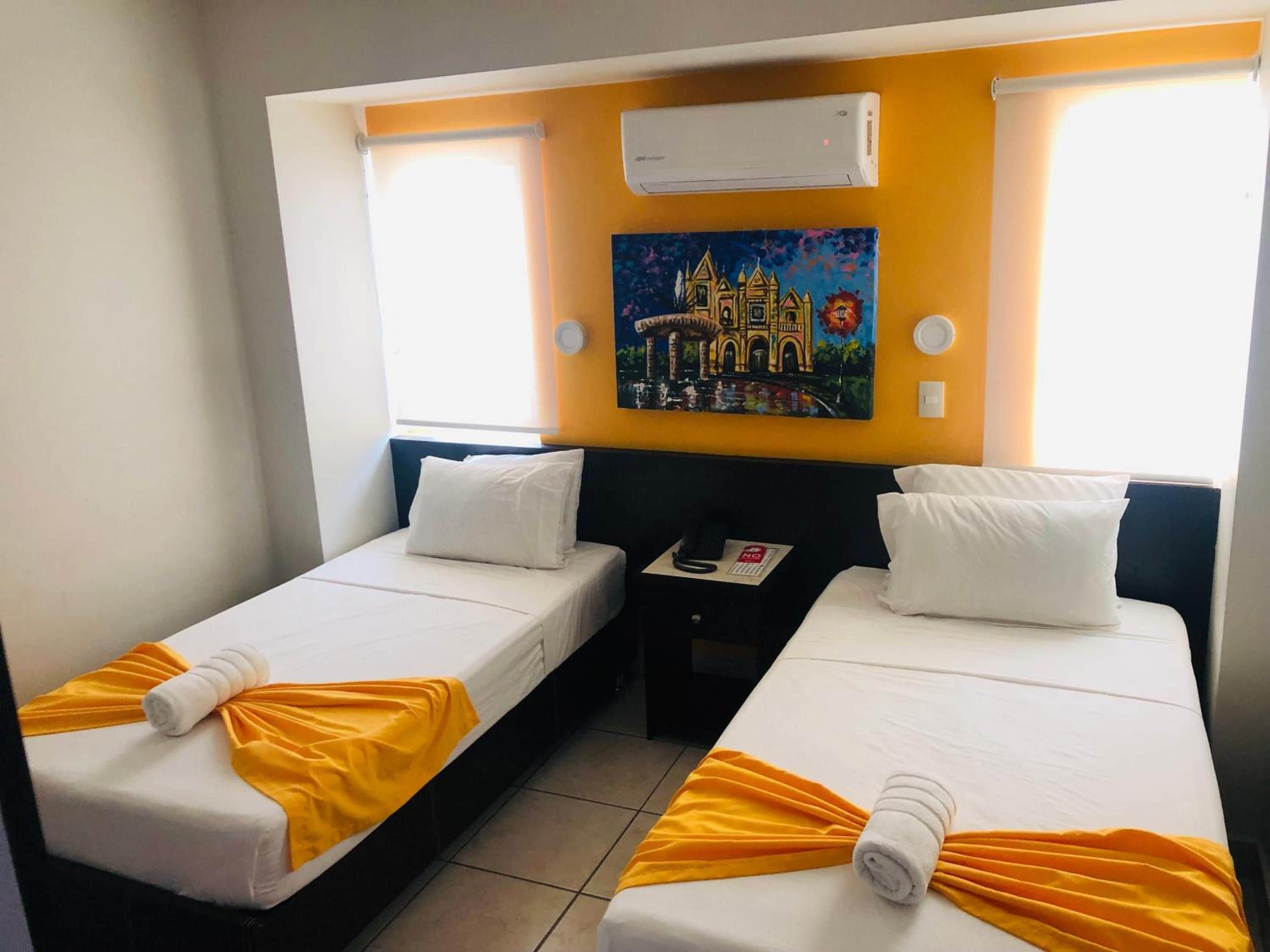 Quarto de hotel com duas camas de solteiro, um quadro de um edifício na parede e um aparelho de ar condicionado sobre as camas.