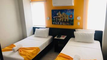 Quarto de hotel com duas camas de solteiro, um quadro de um edifício na parede e um aparelho de ar condicionado sobre as camas.