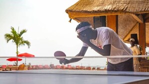 Uma pessoa jogando tênis de mesa em uma quadra externa, com uma estrutura de telhado de palha ao fundo.