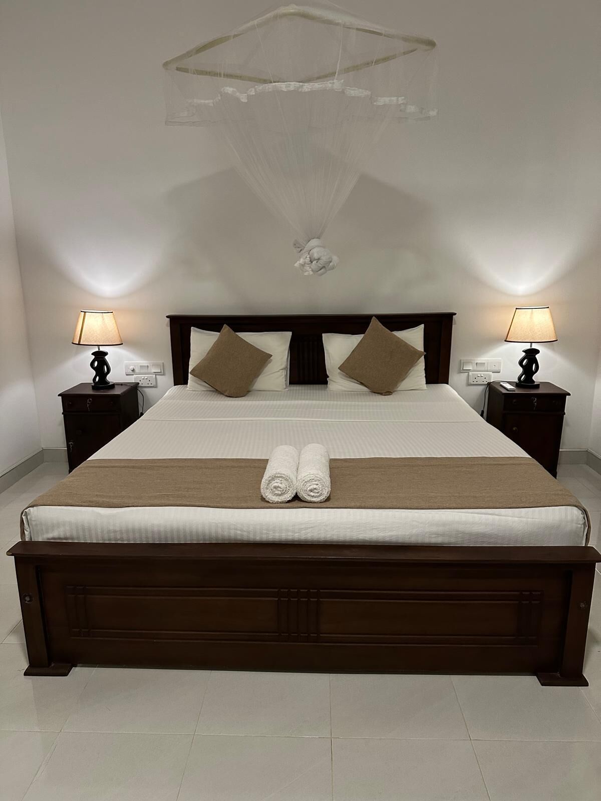 Uma cama grande com estrutura de madeira, duas mesas de cabeceira com abajures e um detalhe decorativo no teto.