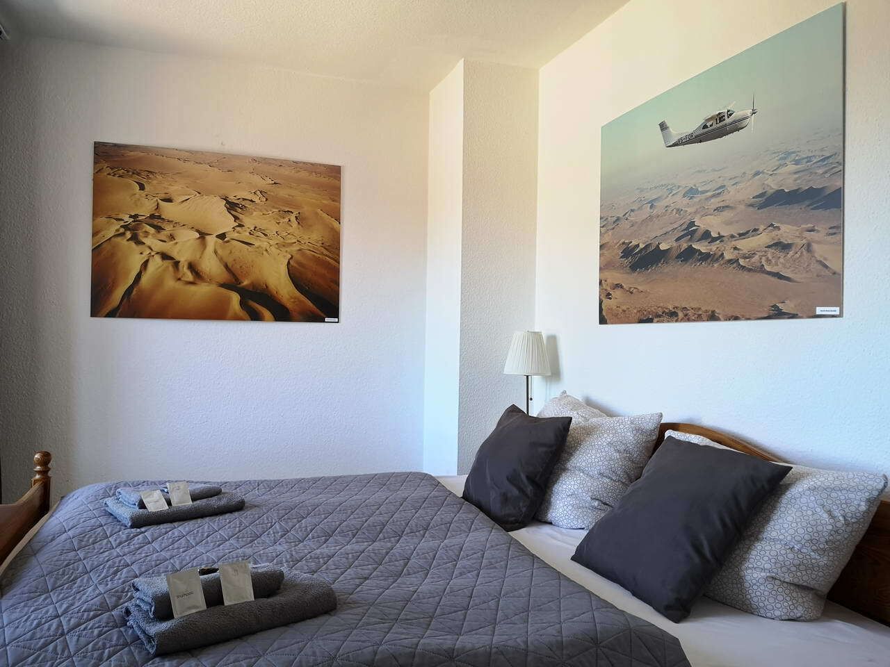 Cama bem arrumada com manta acolchoada, luminária de cabeceira e duas pinturas penduradas na parede retratando paisagens desérticas.