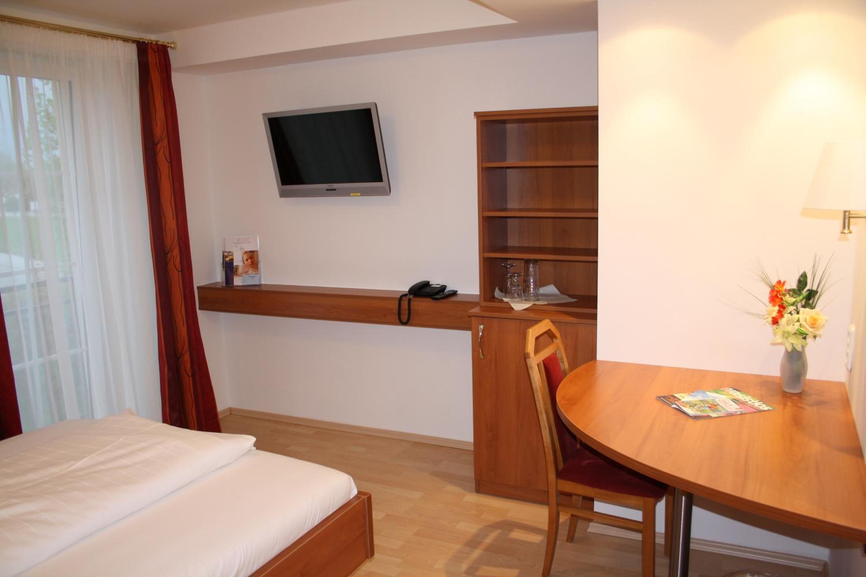 Quarto de hotel com cama, escrivaninha com cadeira, televisão na parede, estante de livros e um vaso com flores na escrivaninha.