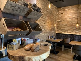 Um ambiente rústico de padaria com prateleiras de madeira exibindo pão e cestos, uma mesa de madeira com pães e uma área de estar com banco e mesa.