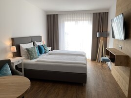 Quarto de hotel moderno com uma cama grande, mesas de cabeceira, escrivaninha, cadeira, televisão e uma janela ampla com cortinas.