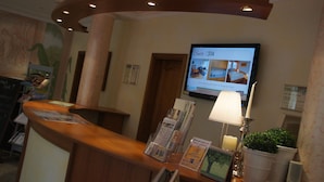 Uma área de recepção de hotel com um balcão curvo, uma TV de tela plana exibindo imagens de quartos, uma luminária, uma vela e um pequeno vaso com planta.