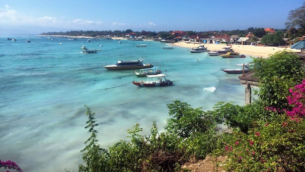 Cena costeira com barcos ancorados perto de uma praia arenosa, cercada por vegetação e edifícios.