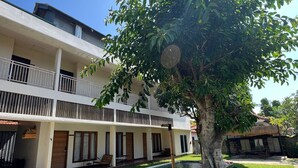 Um edifício de dois andares com piscina, uma árvore e uma construção com telhado de palha.