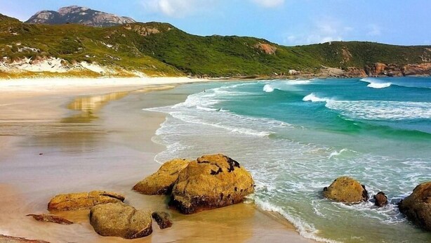 Uma praia com águas turquesa cristalinas, grandes rochas e colinas verdes ao fundo.