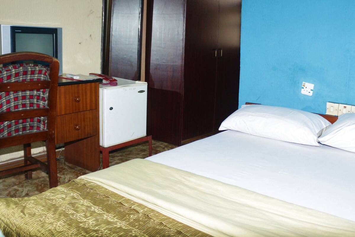 Um quarto com uma cama, uma cadeira de madeira, uma cômoda pequena branca e um guarda-roupa.