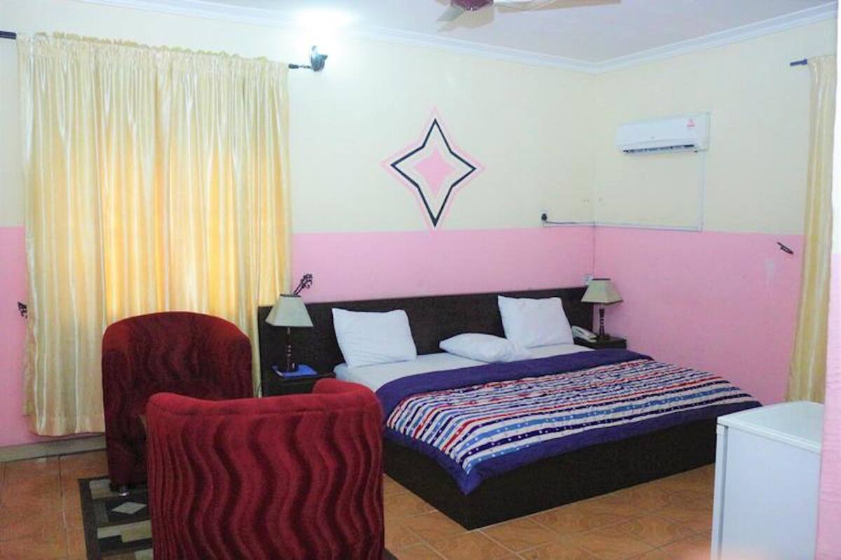 Quarto com cama, mesa de cabeceira, luminária, cadeira e janela com cortinas.