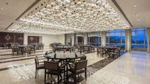 Uma sala de conferências ampla, com várias mesas e cadeiras, teto alto com diversas luminárias, e grandes janelas que oferecem vista para o exterior.