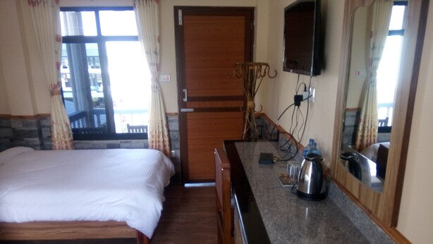 Quarto de hotel com uma cama, mesa de cabeceira de madeira, tapete vermelho, área de cozinha com chaleira e televisão fixada na parede.